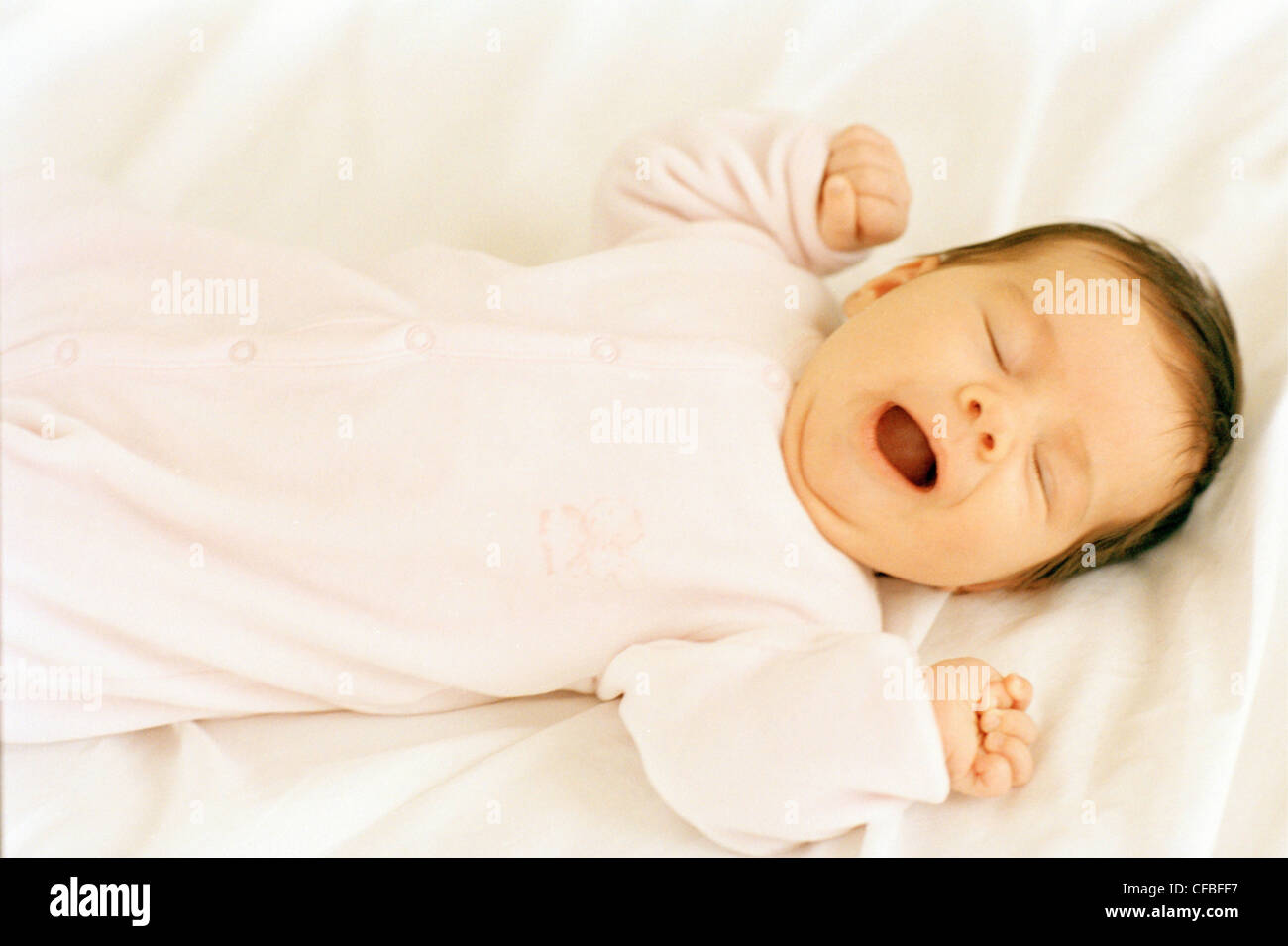 Baby with dark brown hair in pink babygro lying on back on covers eyes ...