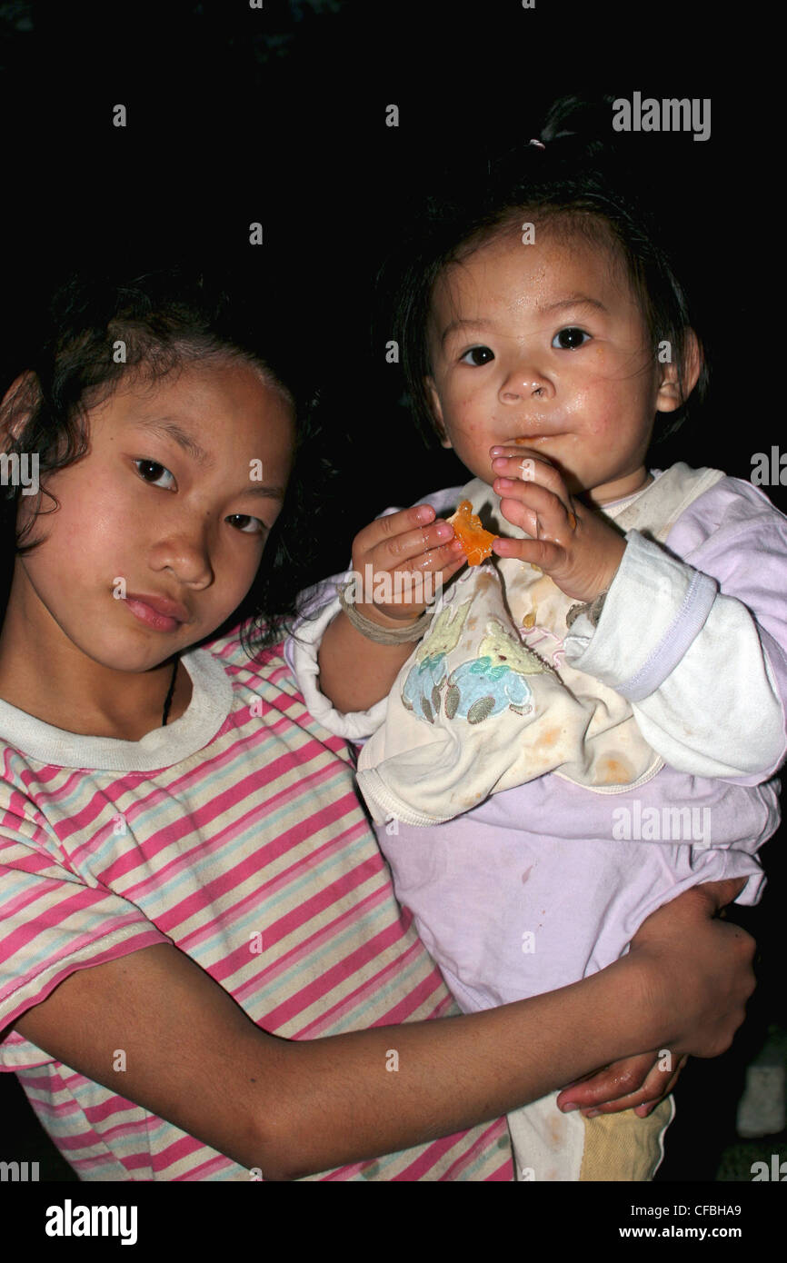 A young Asian girl is holding an infant who is eating fruit in Pai ...