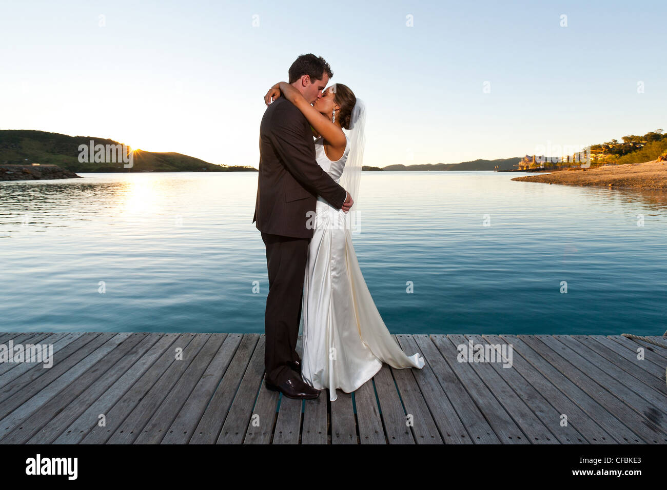 Newlyweds kissing on a jetty at sunset with beautiful water behind ...