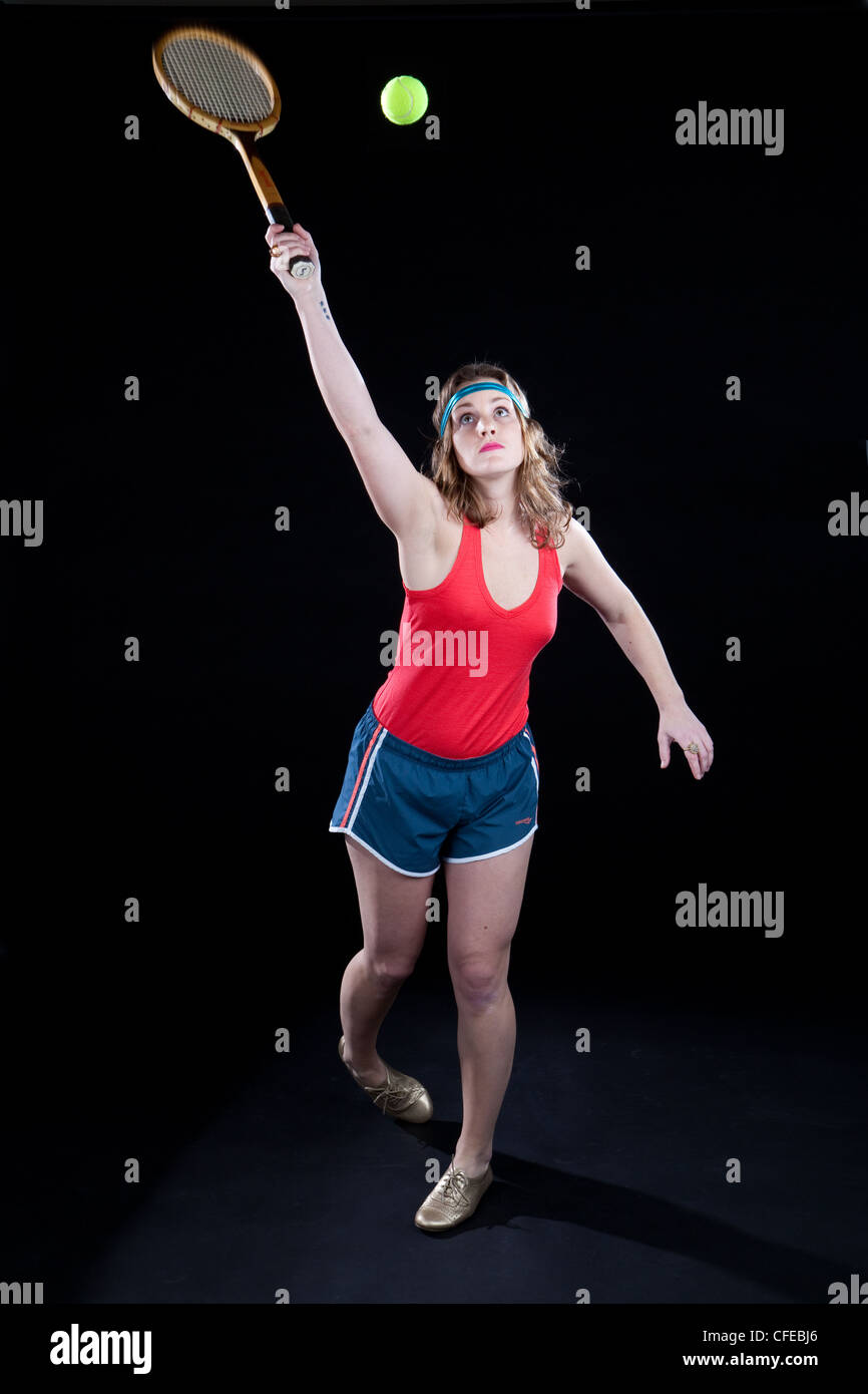 Awkward retro athlete serves a tennis ball in front of a black backdrop ...