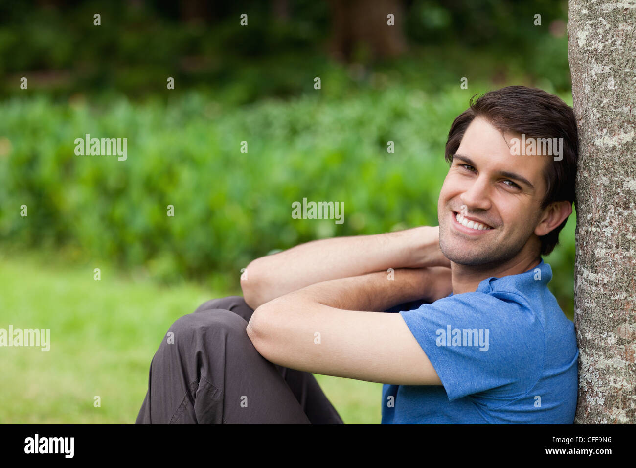 Young smiling man sitting against a tree with his arms resting on his ...