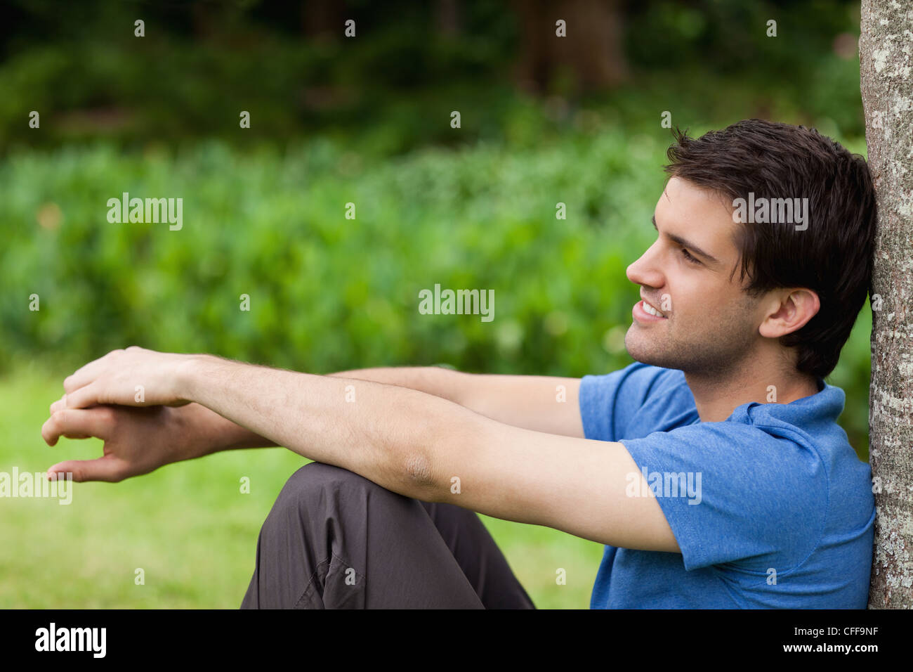 Young man leaning against a tree while placing his arms on his knees ...
