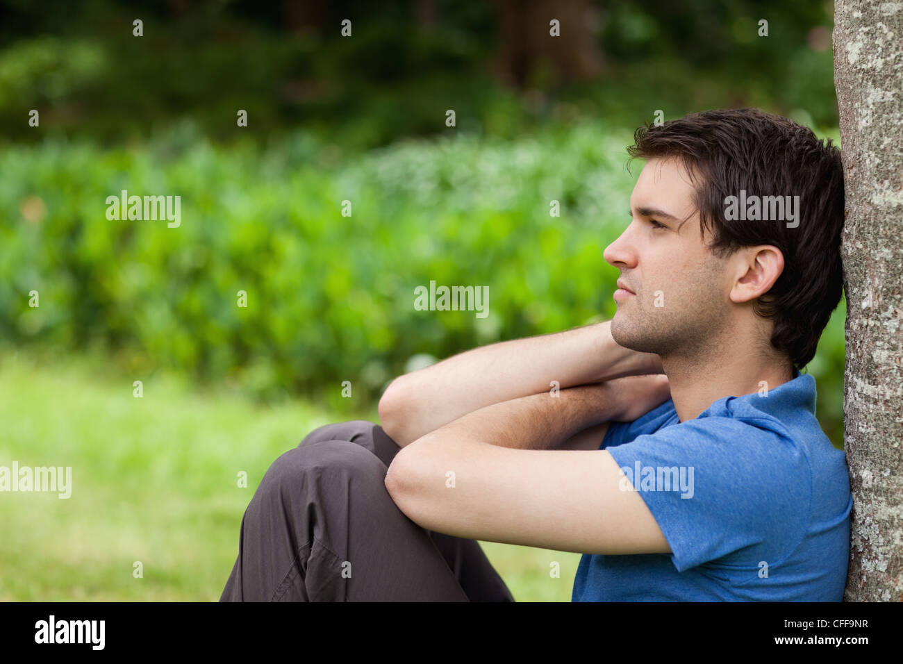 Serious young man sitting against a tree while placing his hands on his ...