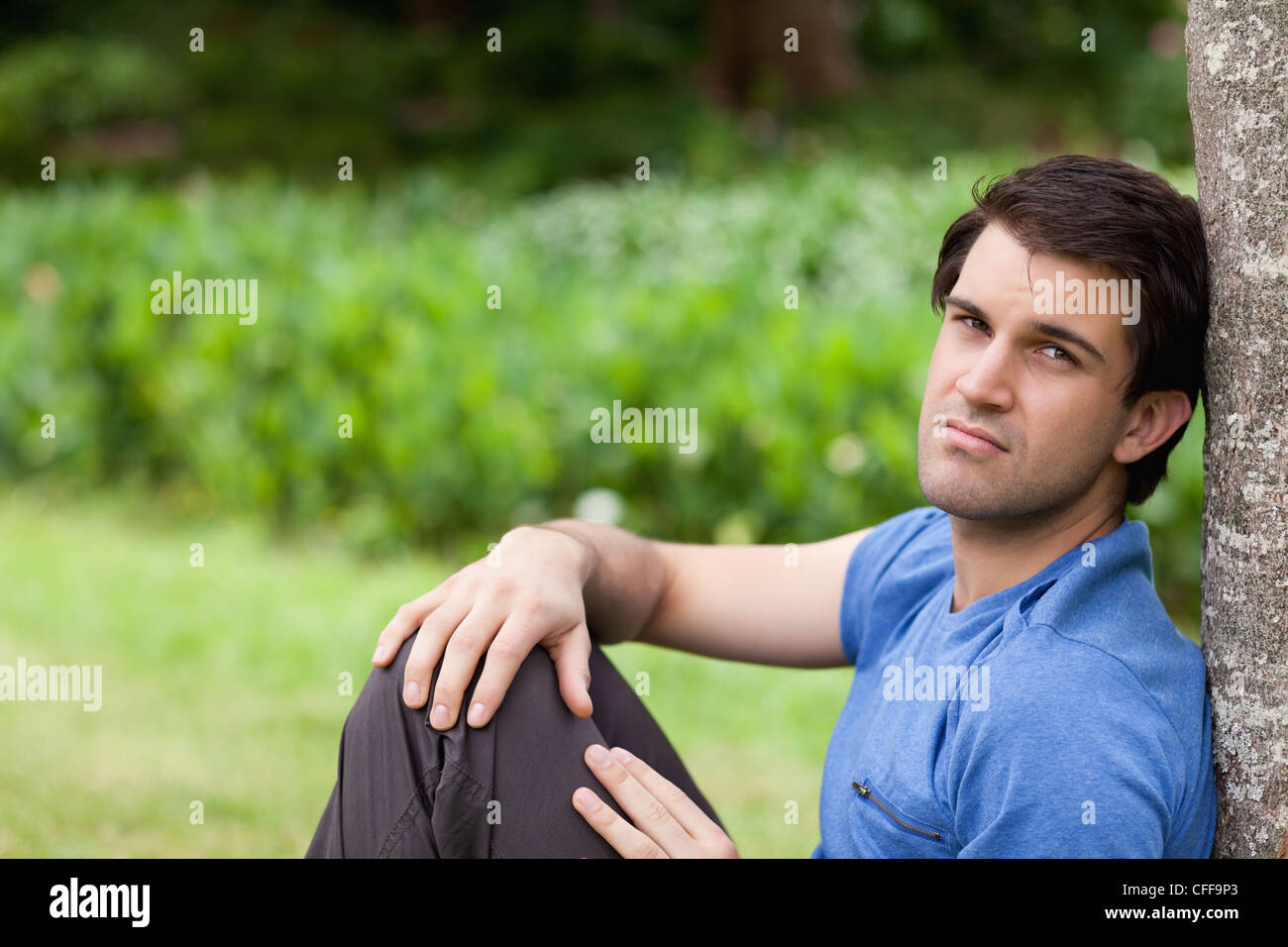 Serious young man leaning against a tree while looking at the camera ...