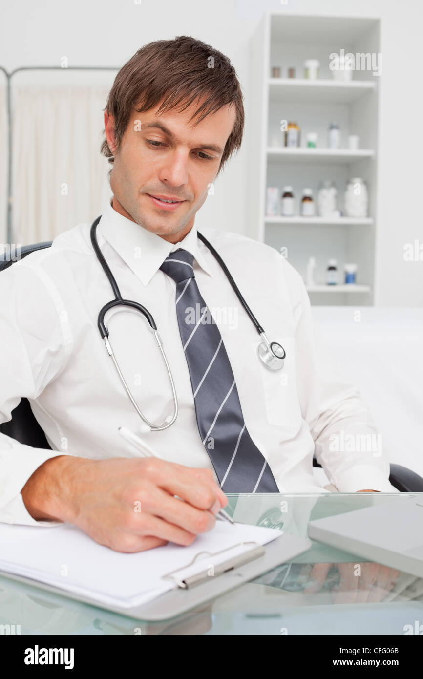 Serious doctor writing on his clipboard while sitting in his office ...