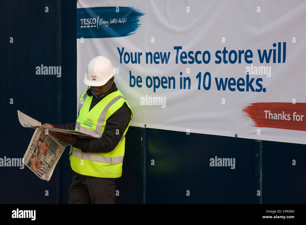 Builder reading the newspaper next to sign saying, "The new Tesco store ...