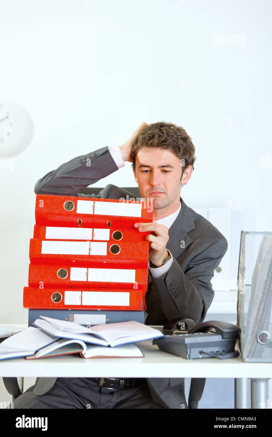Confused modern businessman sitting at office desk with pile of folders ...