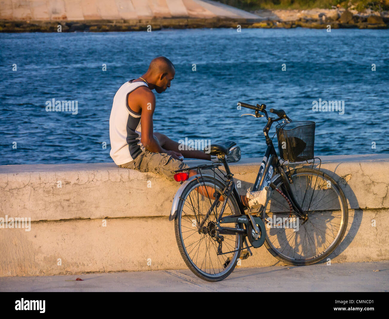 A dark skinned Cuban male rests by sitting on the wall of the Malecón ...