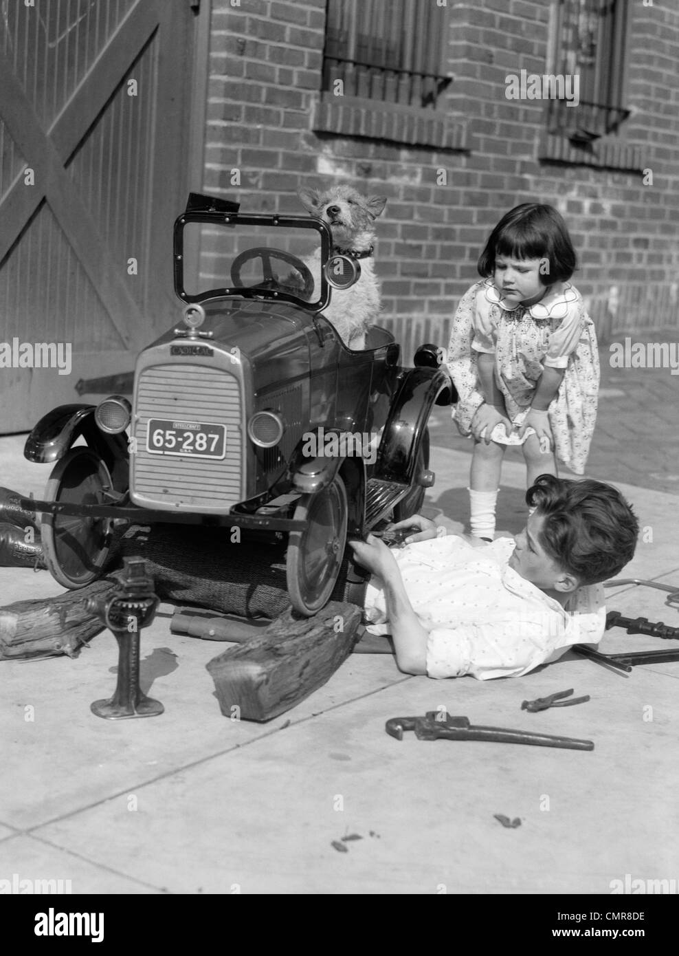 1920s LITTLE GIRL LEANING DOWN WATCHING BOY WORKING UNDER TOY CAR WITH ...
