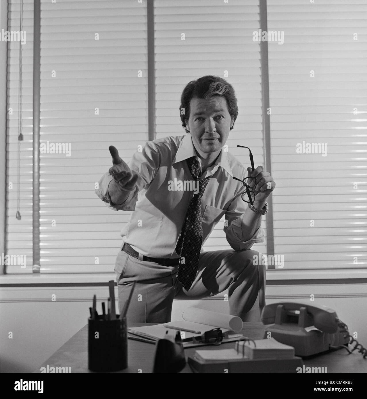 1970s BUSINESSMAN STANDING BEHIND DESK ARM RAISED IN PLEADING GESTURE ...