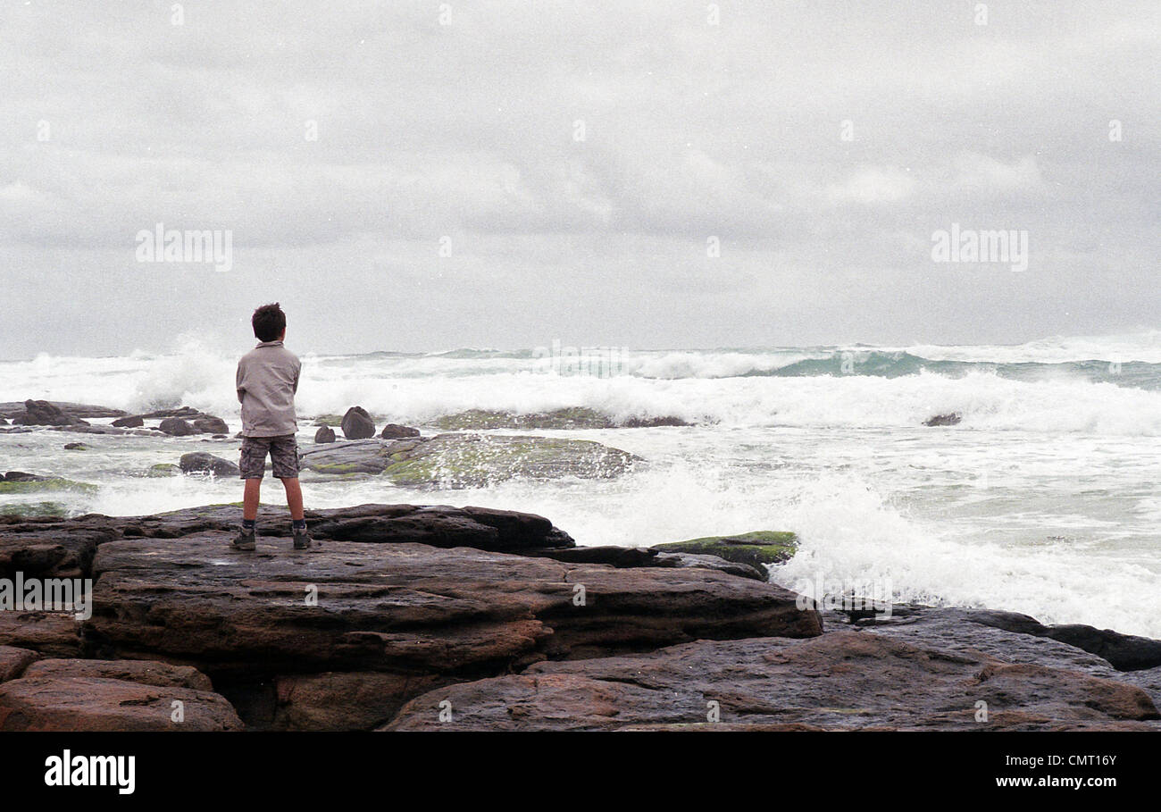 A boy stands on rocks facing a raging sea and foreboding clouds Stock ...