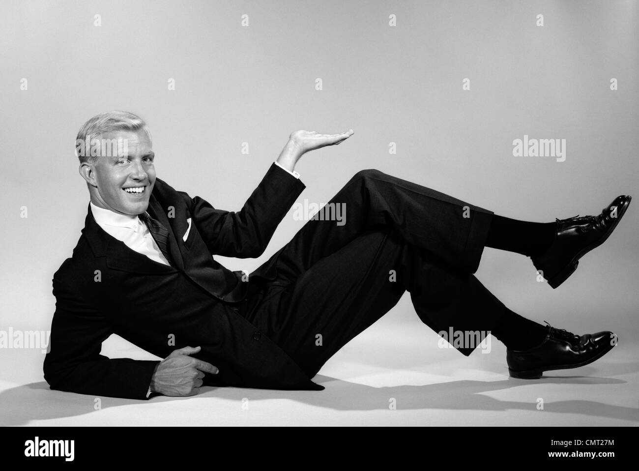 1960s SMILING BUSINESSMAN RECLINING PROPPED ON ELBOW HOLDING HAND UP IN ...