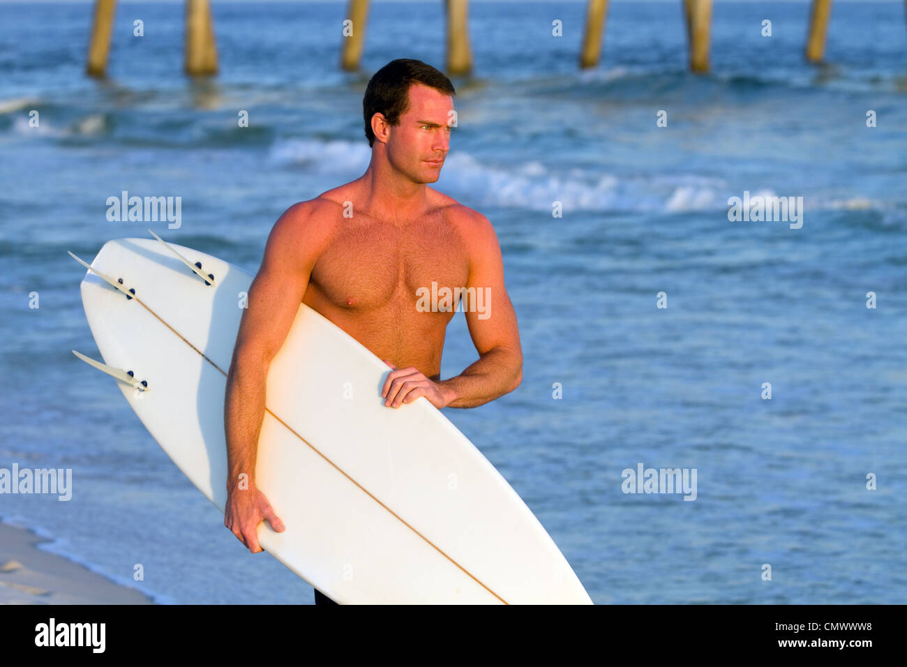 Young athletic adult male surfer carries his surfboard along the beach ...