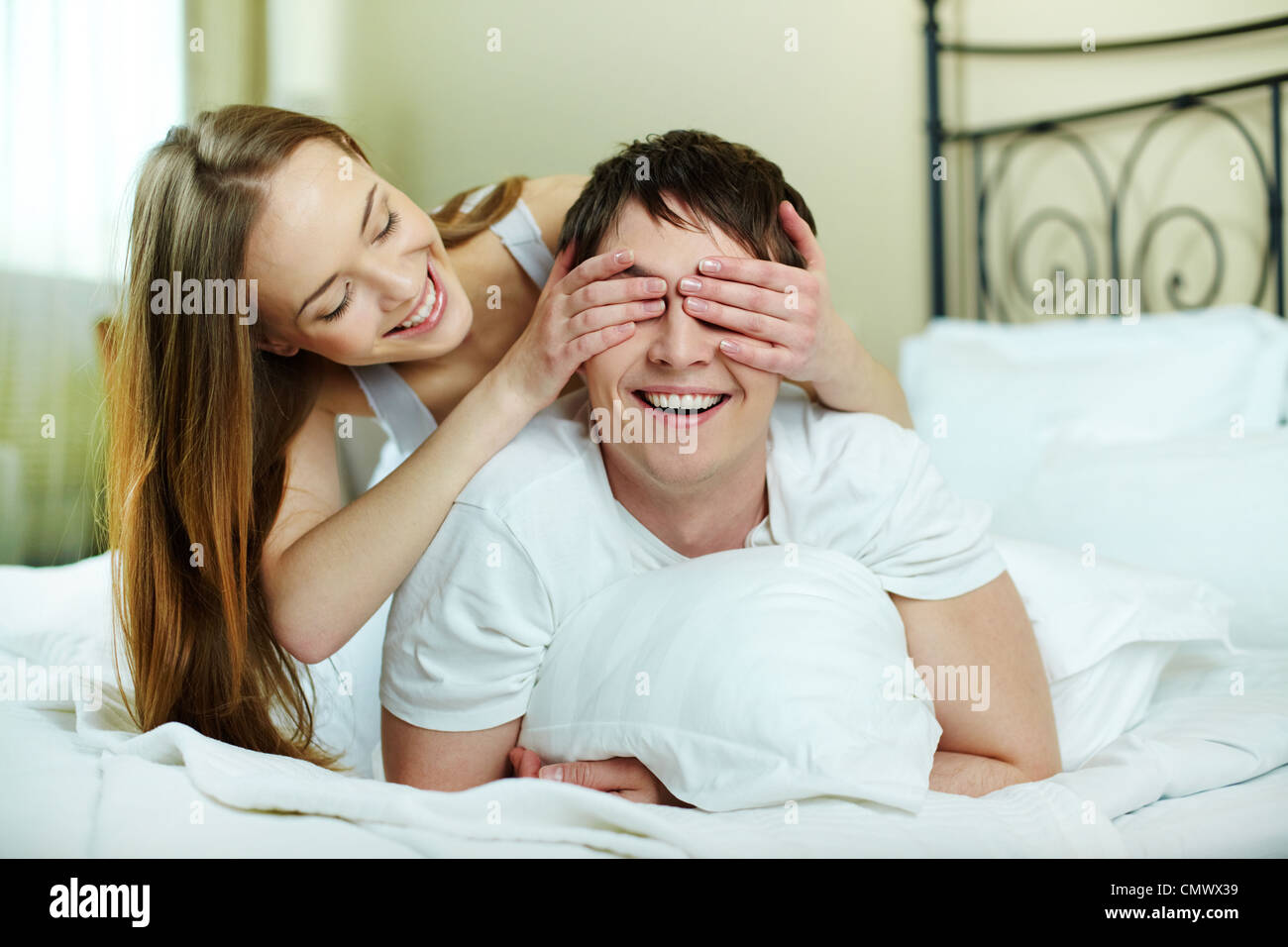 Happy girl closing her boyfriend eyes by hands in bed Stock Photo - Alamy