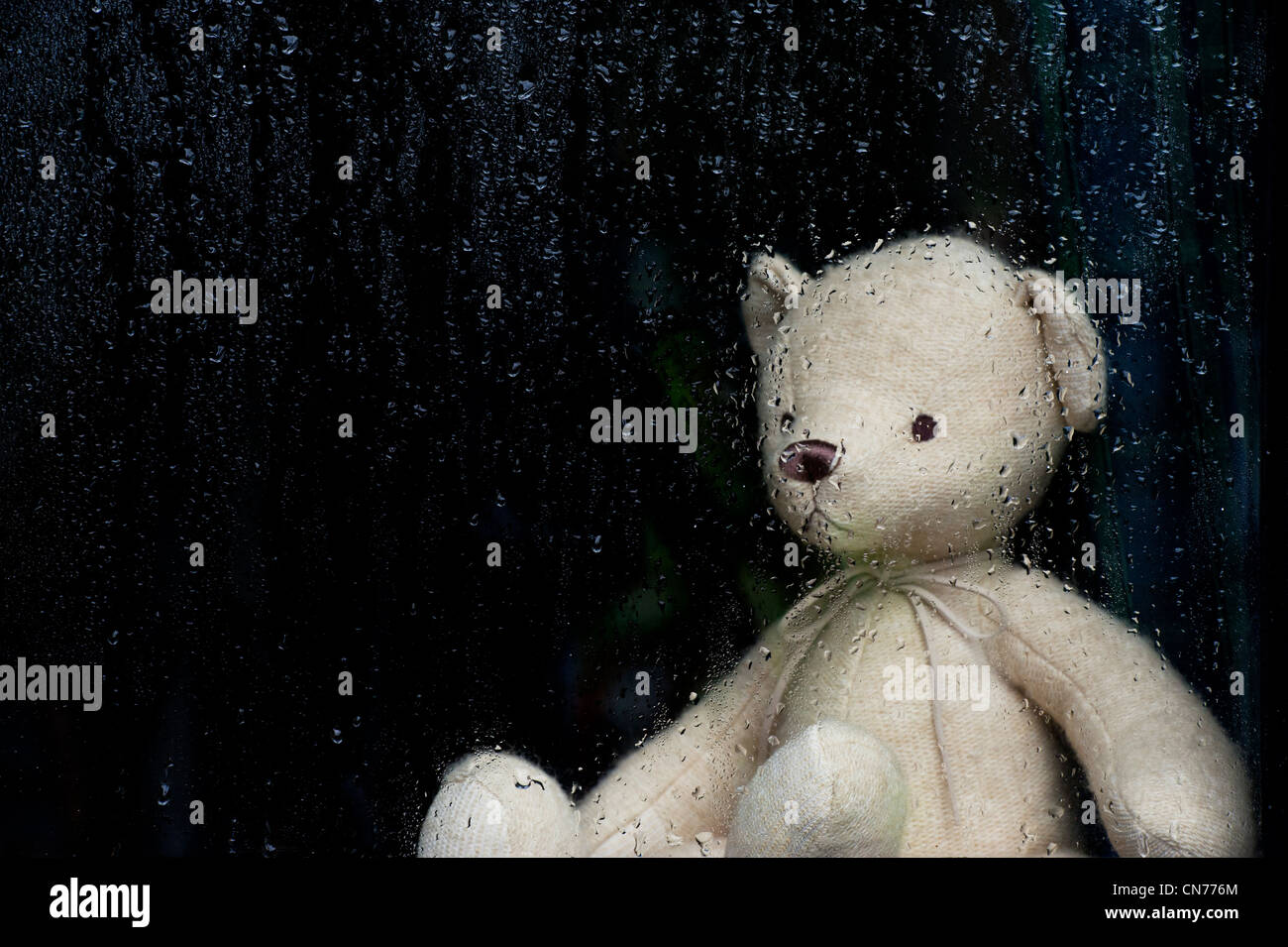 Sad Teddy bear looking through a window covered in rain drops. Still ...