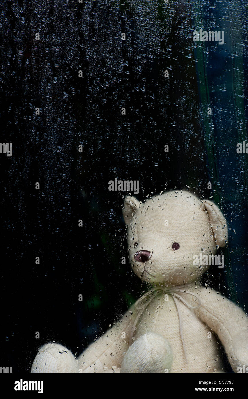 Sad Teddy bear looking through a window covered in rain drops. Still ...