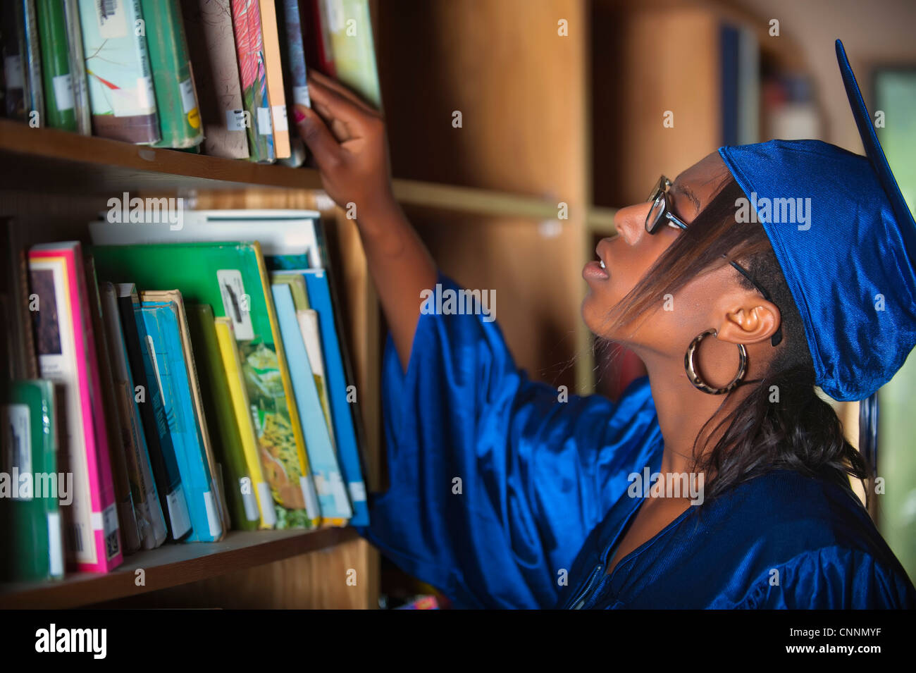 Female student in graduation outfit looking at bookshelf at library ...