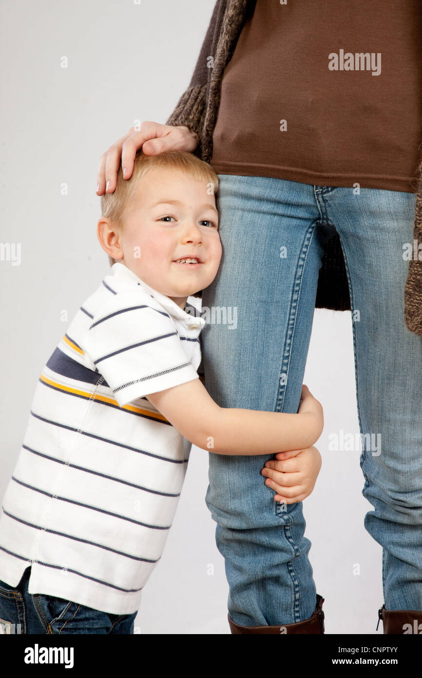 Cute blond boy, tightly holding onto his mothers leg, as she rests her ...