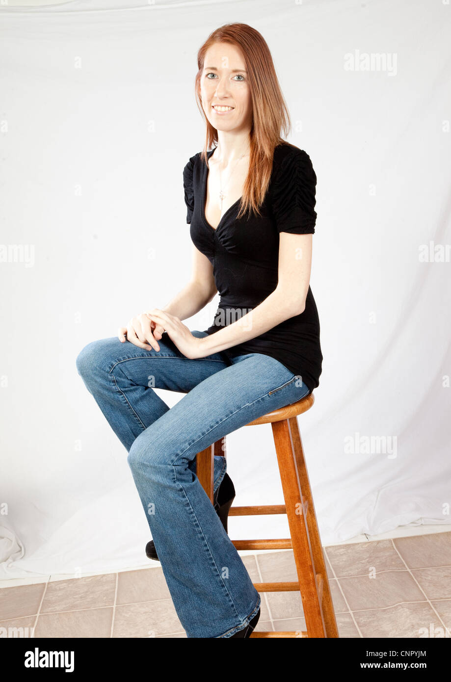 Pretty woman with long hair and wearing blue jeans, sitting on a stool ...