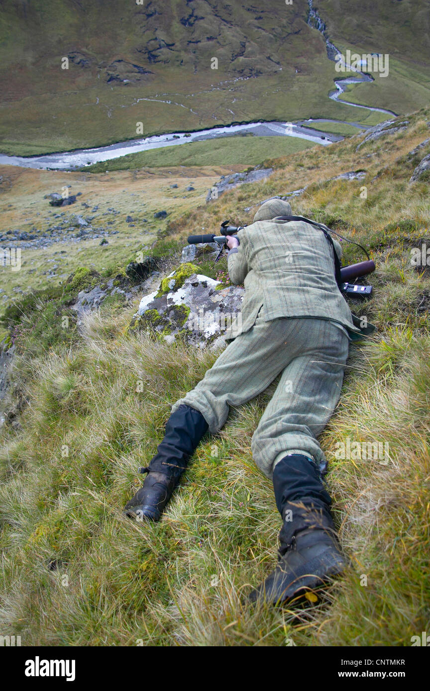 stalker lying on the belly at the edge of a mountain slope aiming down ...