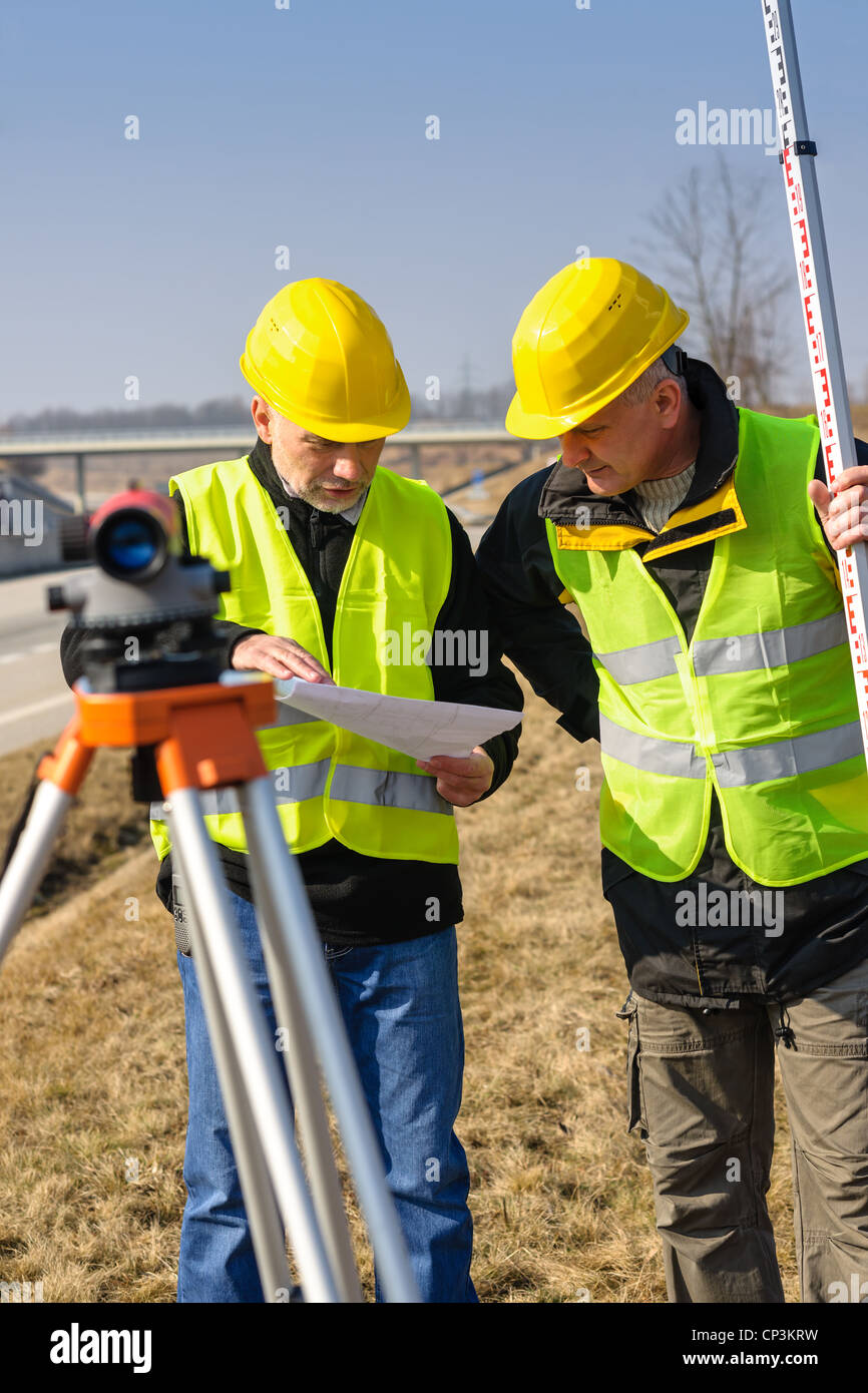Two male geodesist with tacheometer checking plans standing by highway ...