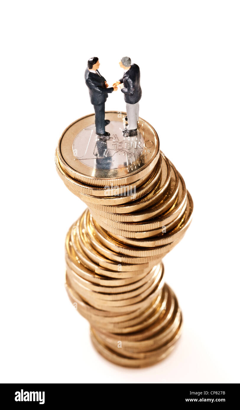 Two business men standing on a wobbly pile of € coins and shake hands ...