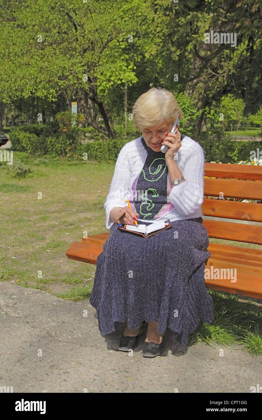 Woman calling by mobile phone and taking notice in a notebook in a park ...