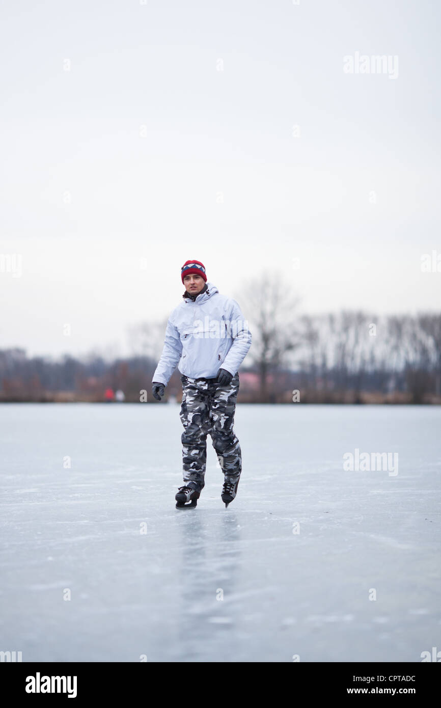 Handsome young man ice skating outdoors on a pond on a cloudy winter ...