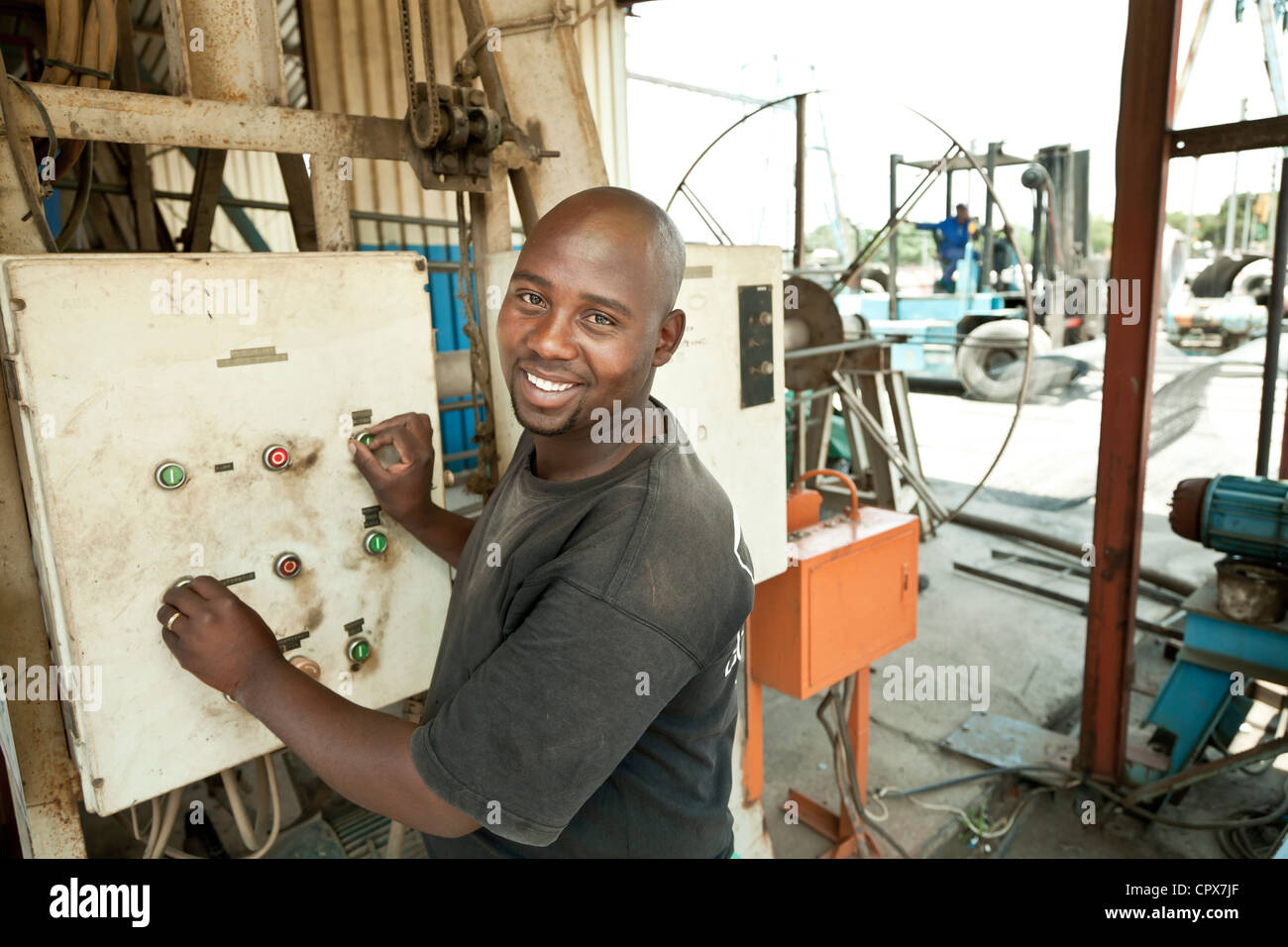 Black factory worker adjusts crane controls while smiling at camera ...