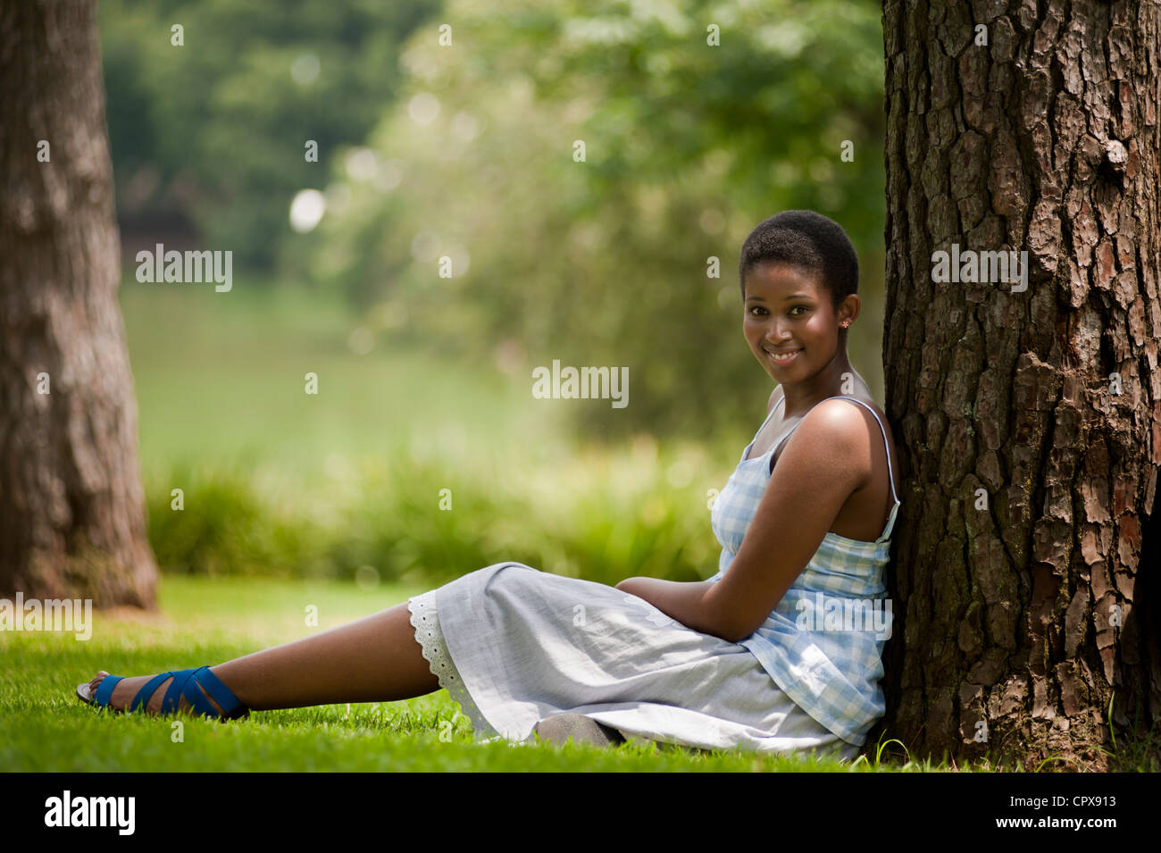 A young African woman sits against a tree in a park, smiling at camera ...