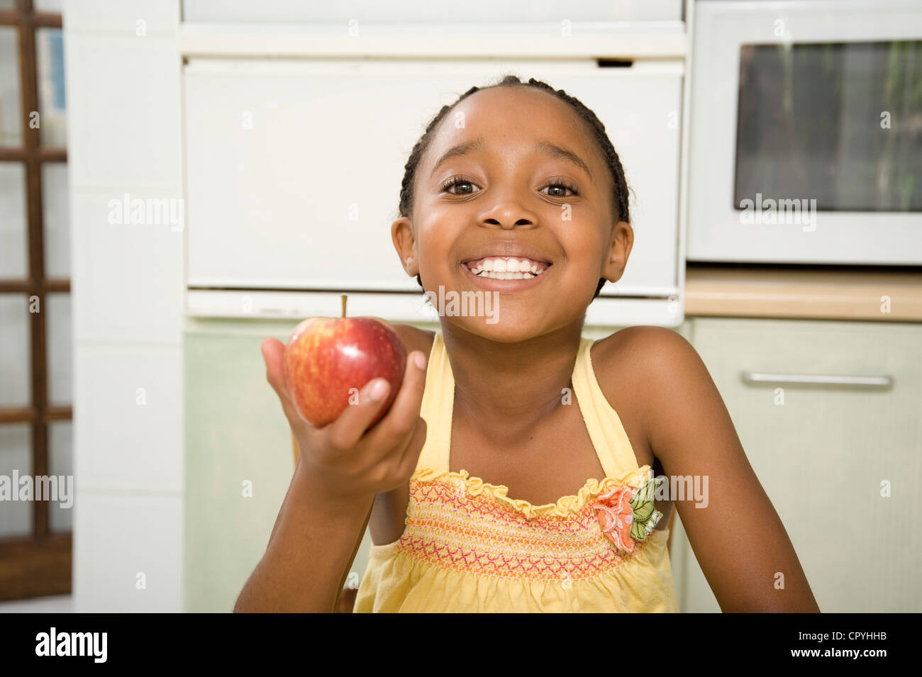 Young African girl sits in a kitchen eating an apple, smiling at camera ...