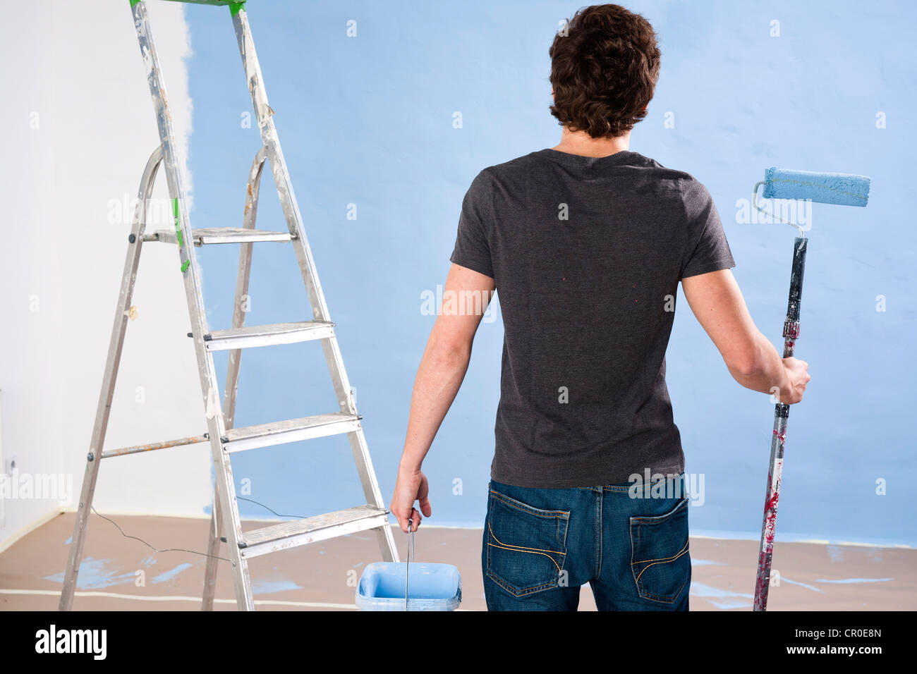 Young man holding a paint roller stands pensively in front of a wall ...