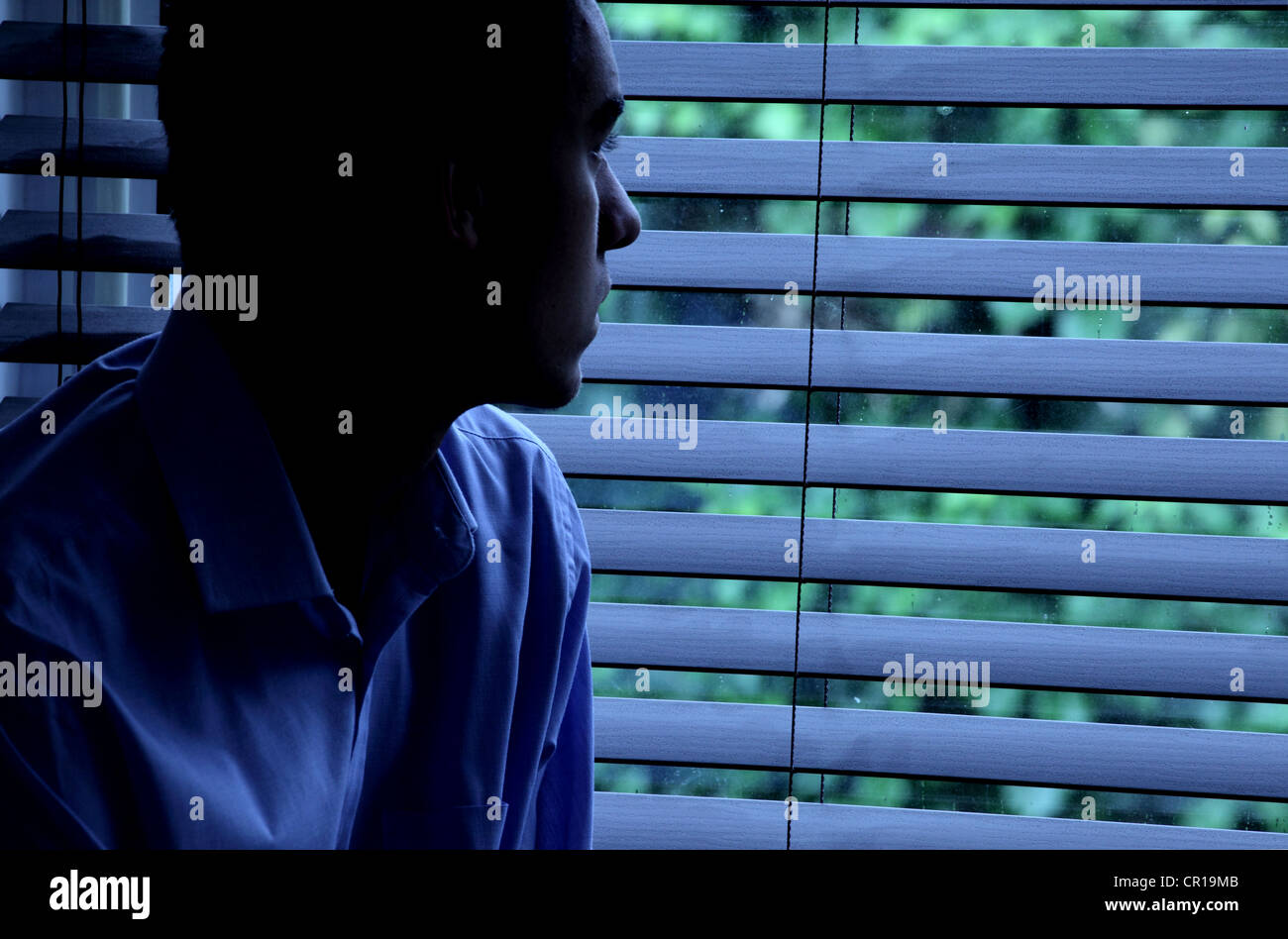 Young man sitting in a dark room looking out through a window blind ...