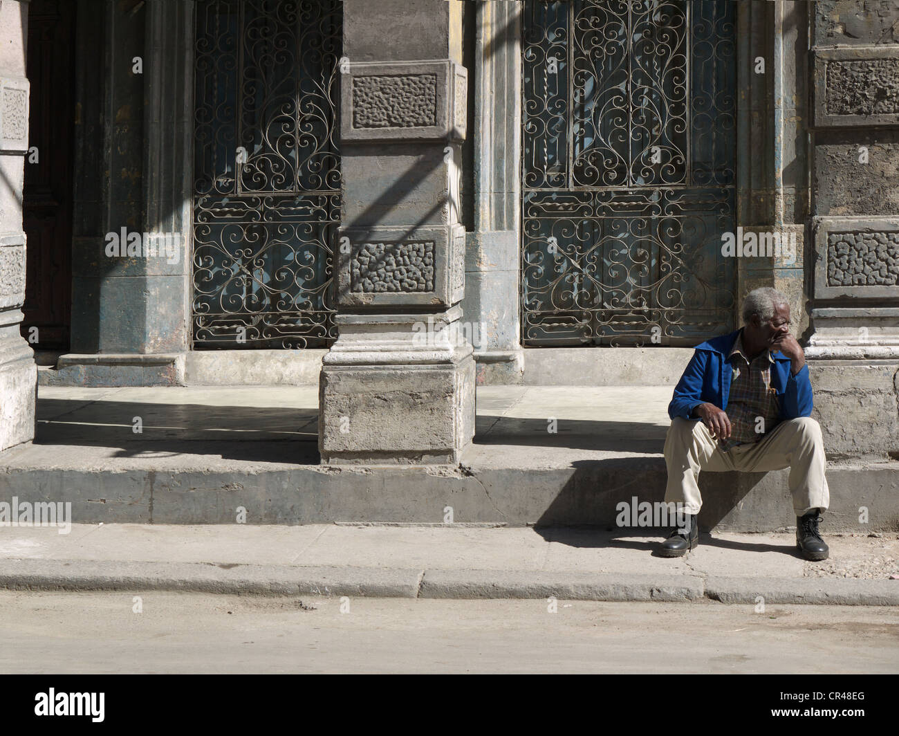 Dark-skinned elderly Cuban man sitting bored on the stairs in front of ...