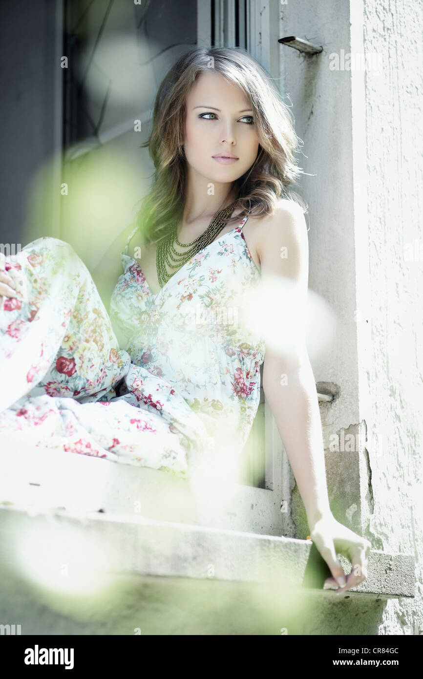 Young woman sitting on a window sill, behind her a broken window pane ...