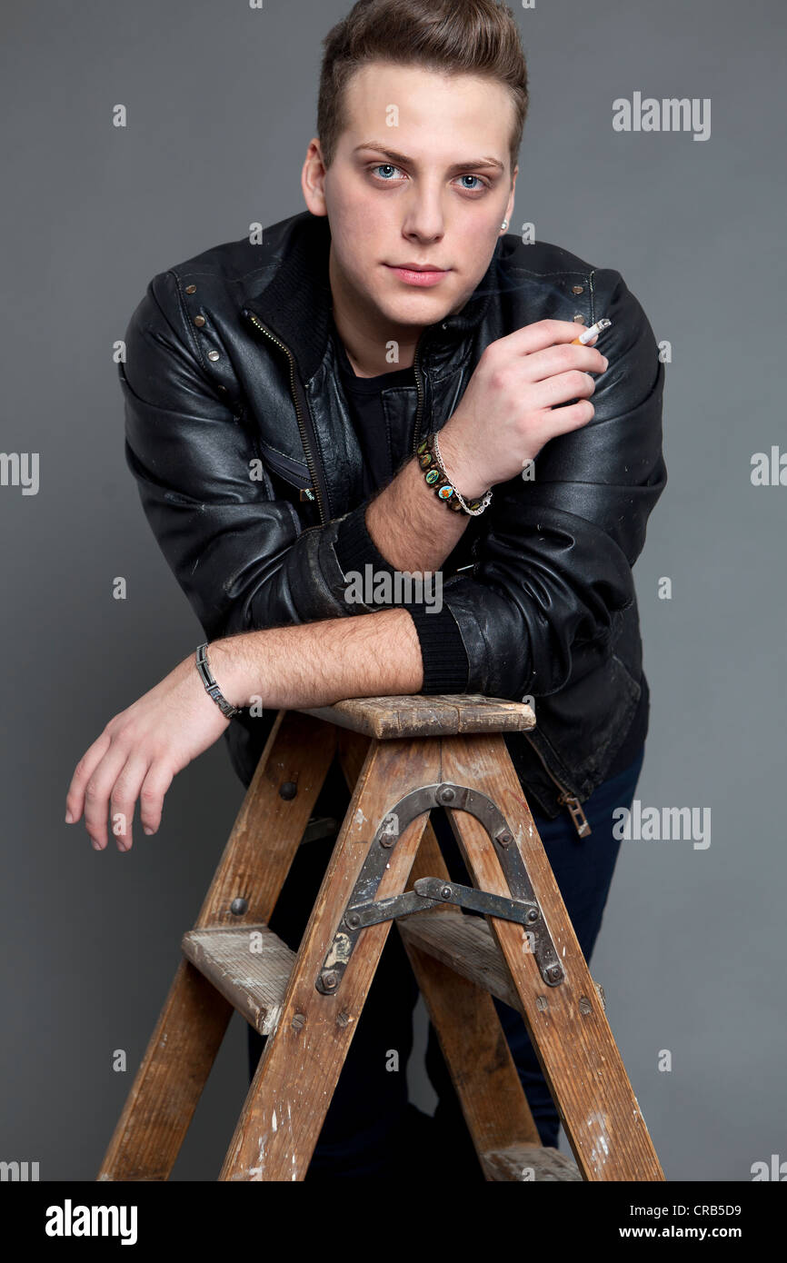 Young man in a leather jacket smoking a cigarette, leaning on a ladder ...