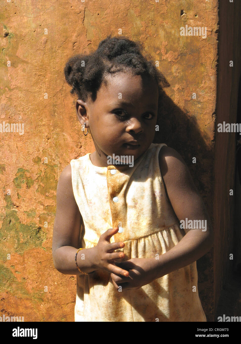 Portrait of a small girl in rural area standing at wall of house ...