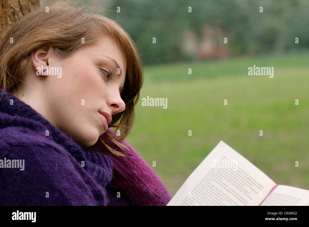 young woman leaning against a tree in a park and reading a book ...