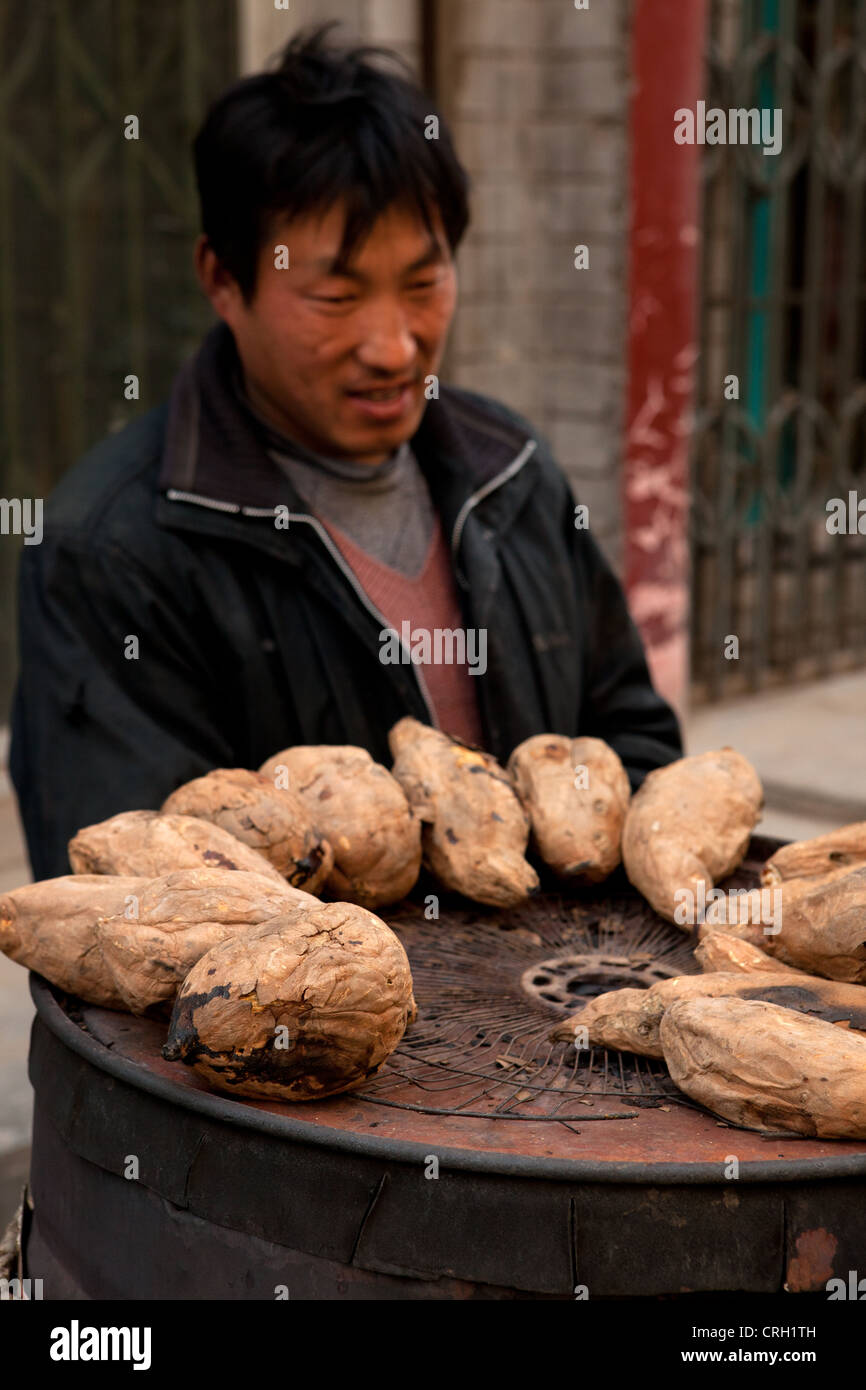 Smiling Chinese man looking at roasted sweet potatoes on pan at street ...
