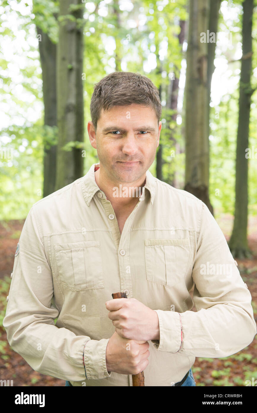 Mid adult man standing in a forest holding a walking stick and looking ...