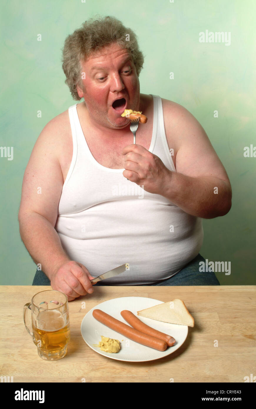 Fat man in undershirt eating from a plate with two sausages, mustard ...