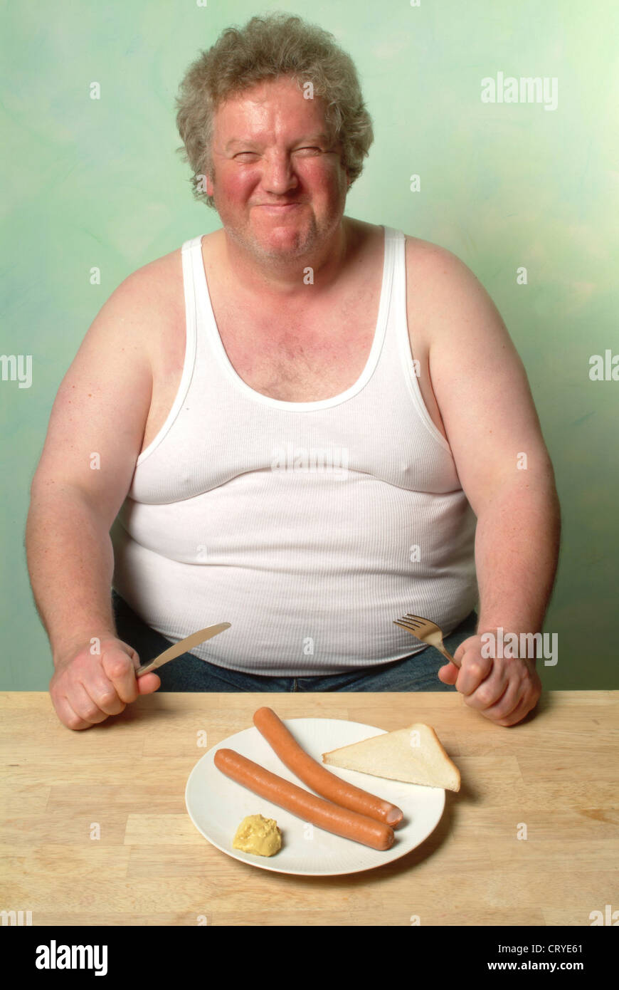 Fat man in undershirt sitting in front of a plate with two sausages and ...