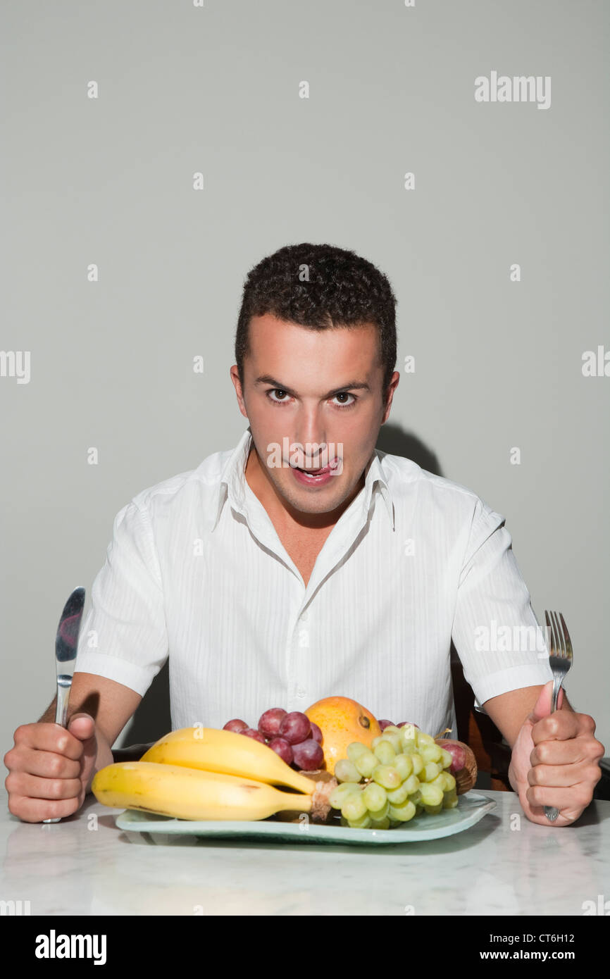 Portrait of young man holding fork and knife with variety fruit on tray ...