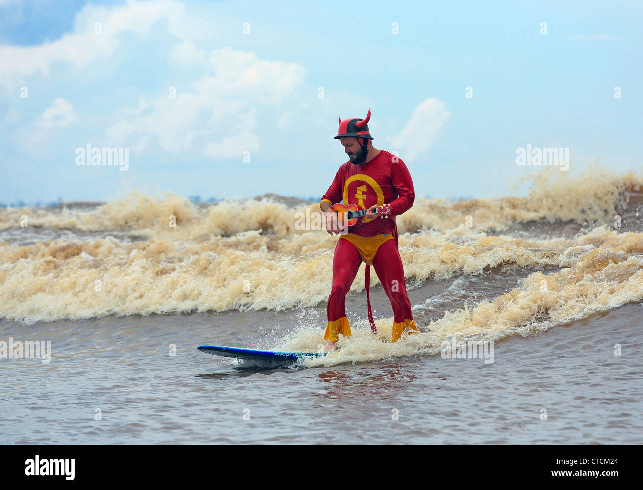 Strange man in red superhero costume river surfing a wave while playing ...
