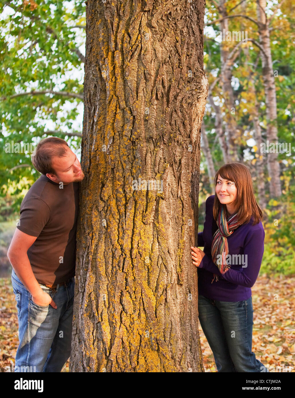 Young Married Couple Peaking At Each Other Around A Tree In A Park In ...