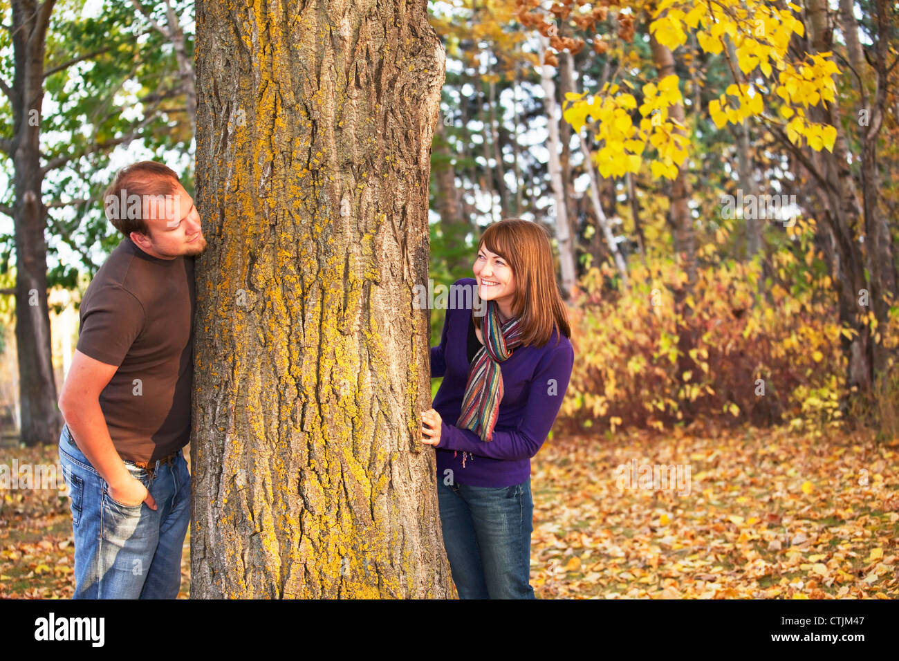 Young Married Couple Peaking At Each Other Around A Tree In A Park In ...