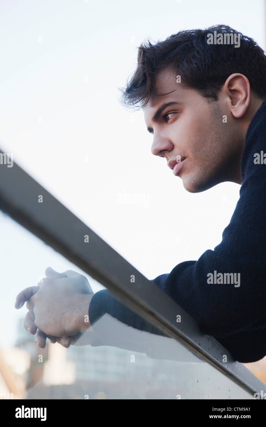 Handsome and thoughtful young man leaning on glass railing of a balcony ...