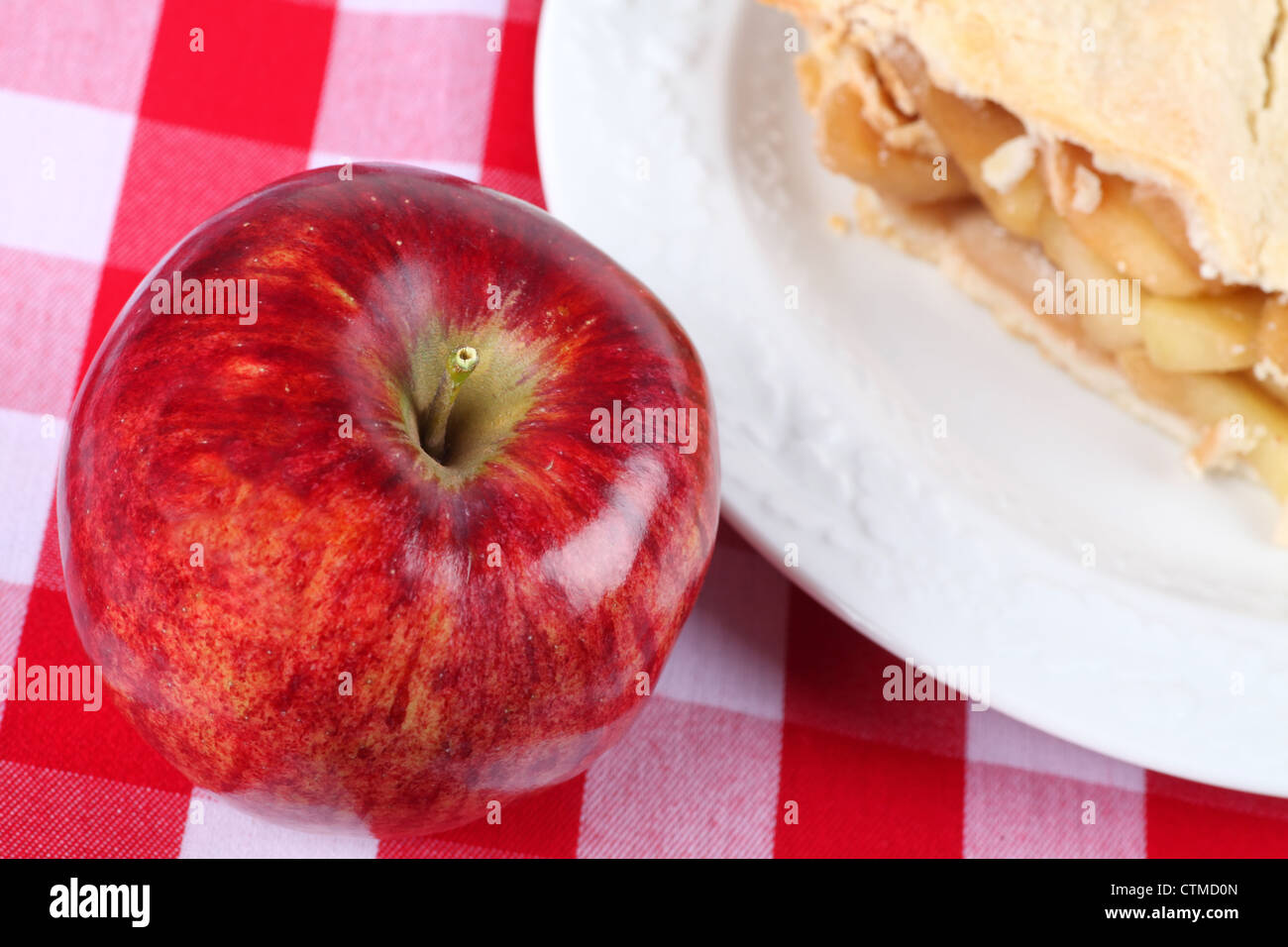A ripe red apple on gingham tablecloth beside a slice of apple pie ...
