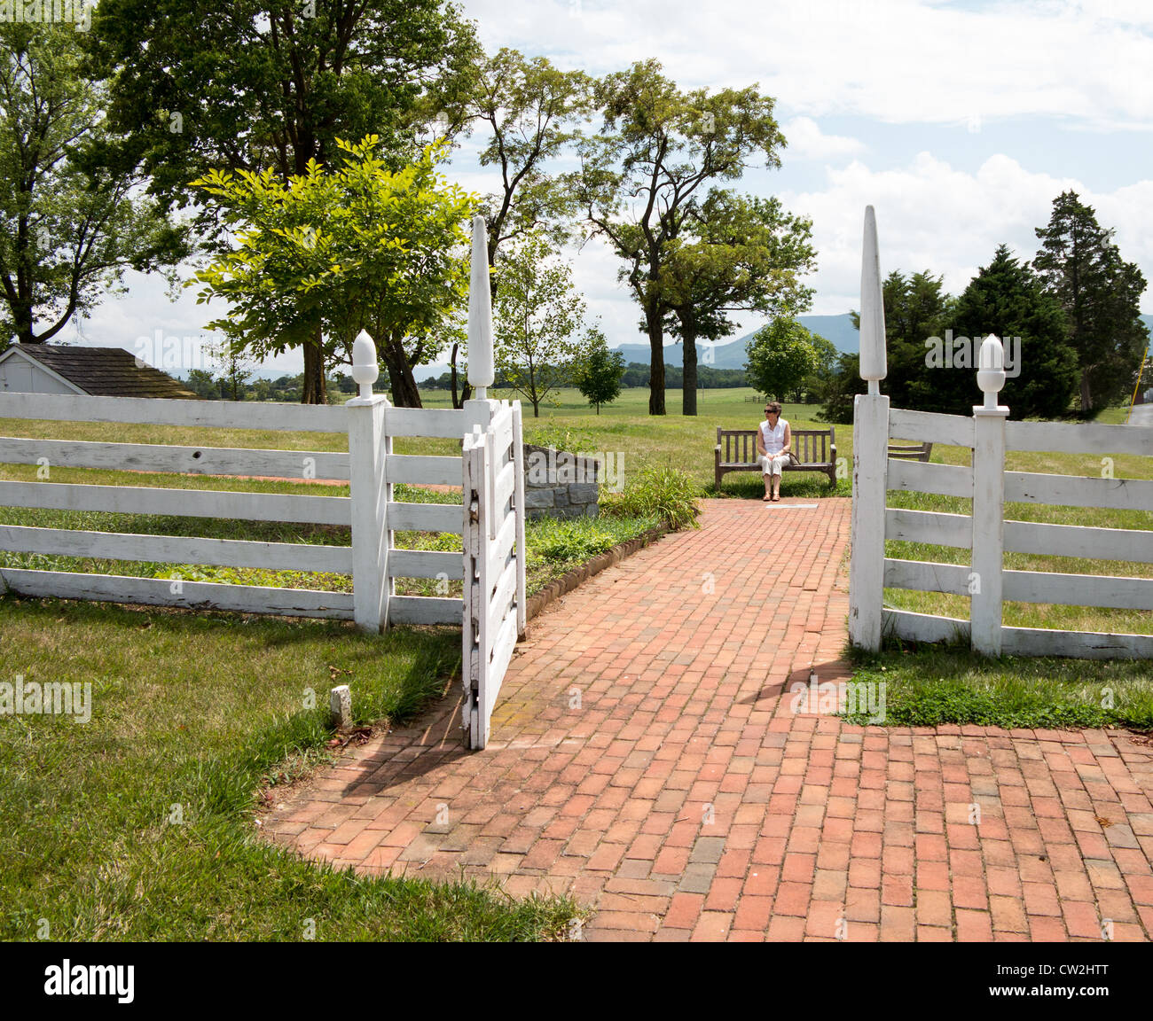 Senior woman sitting on bench on brick path through white picket gate ...
