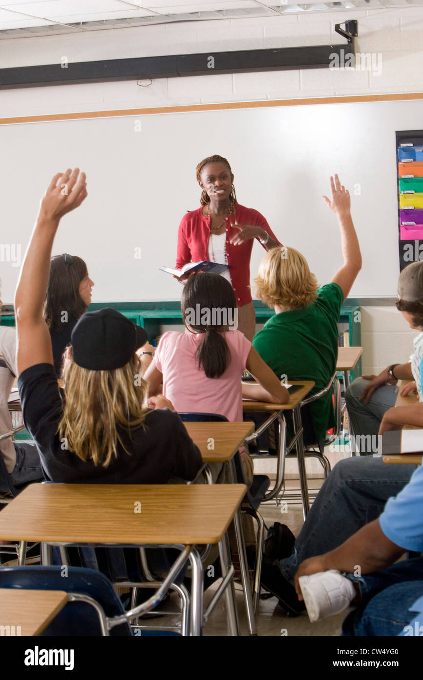 Cheerful female teacher holding book and students raising hands in the ...
